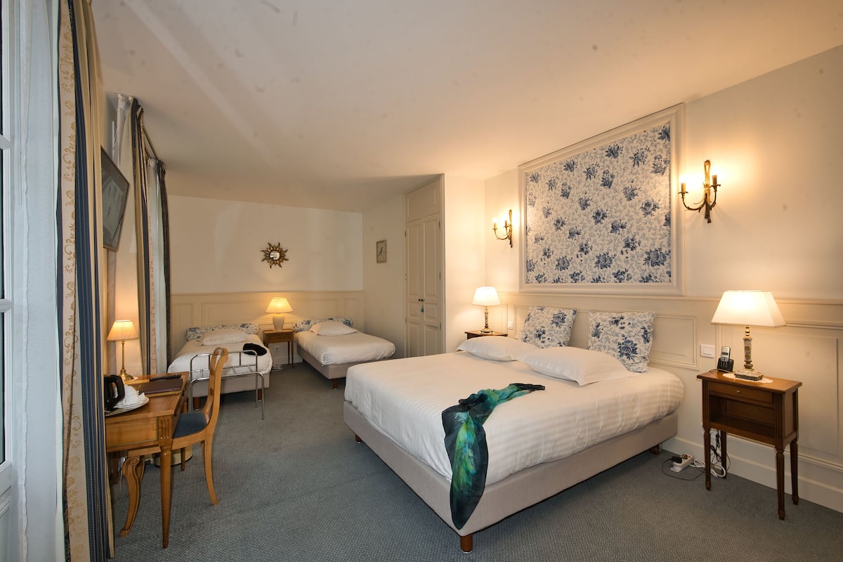 Quarto de hotel com duas camas de solteiro, uma escrivaninha com cadeira, mesa de cabeceira com abajur e um grande painel de parede com padrão floral.