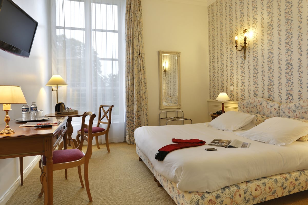 Quarto de hotel com cama, escrivaninha, cadeira, televisão e janela com cortinas.