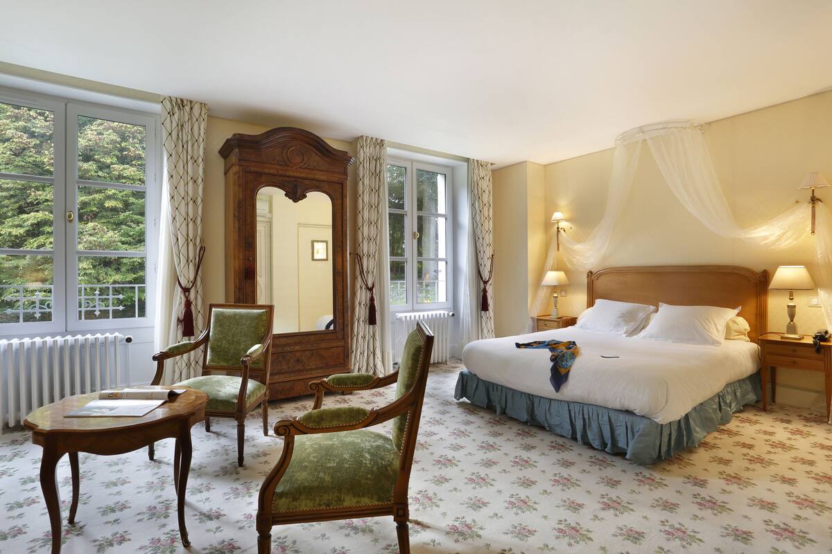 Quarto de hotel com uma cama grande, duas mesas de cabeceira com abajures, uma cadeira, um espelho e uma janela com cortinas.