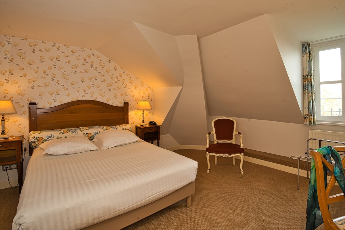 Quarto com uma cama grande, duas luminárias de cabeceira, um painel de cama de madeira, parede com padrão floral, uma cadeira vermelha e uma janela com cortinas.