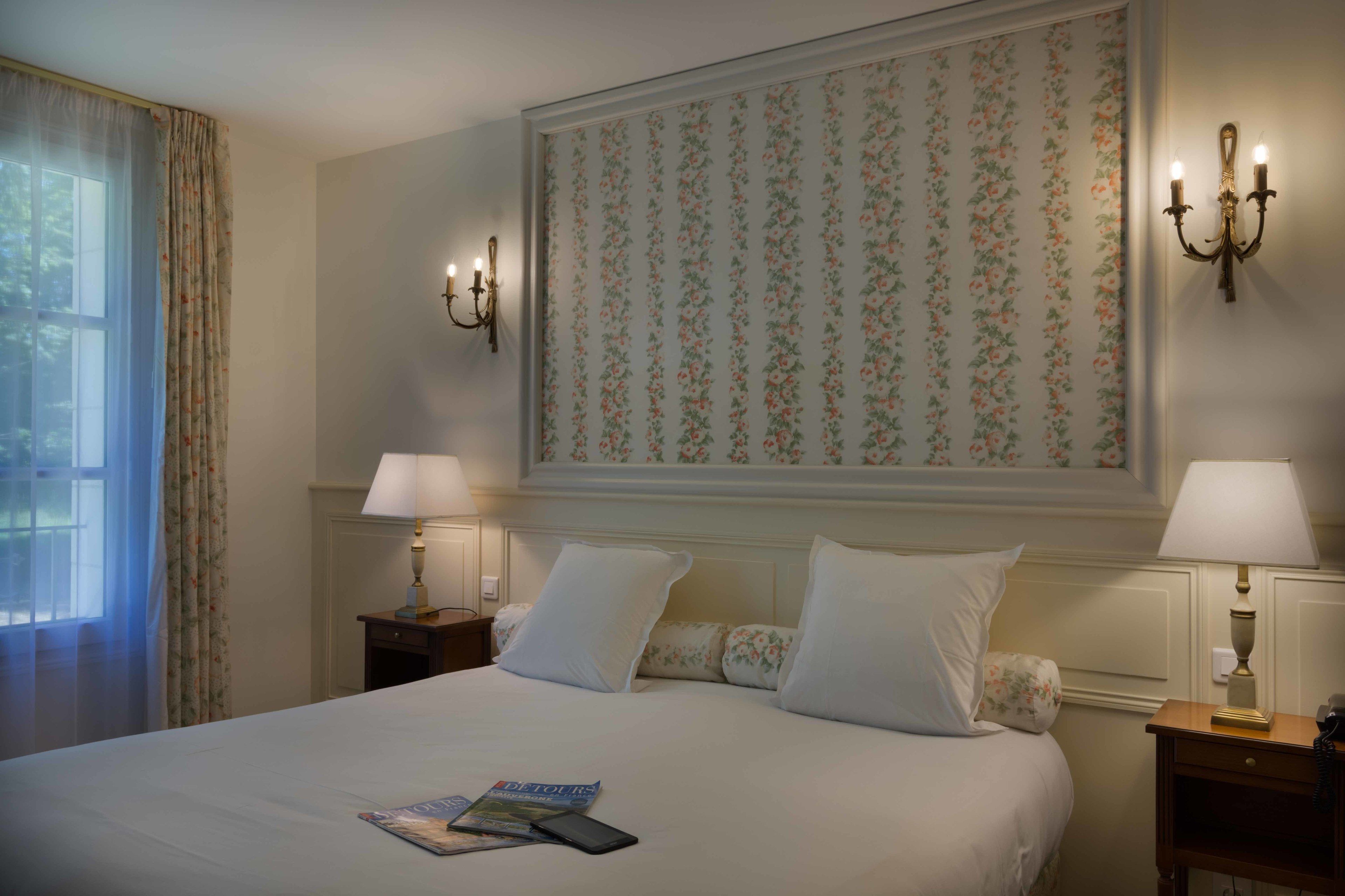 Uma cama bem arrumada com roupa de cama branca, papel de parede floral e duas luminárias fixadas na parede.