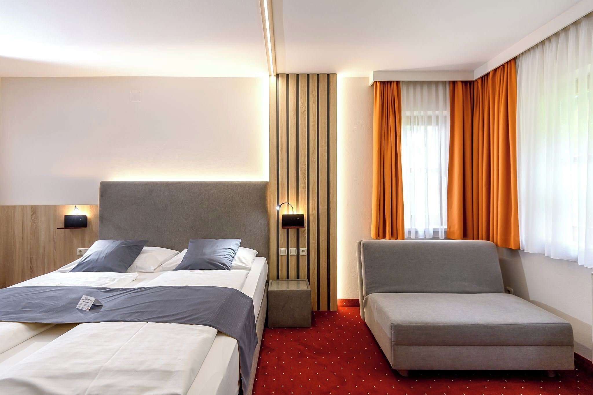 Quarto de hotel com duas camas, um sofá, uma mesa pequena e uma luminária na parede.