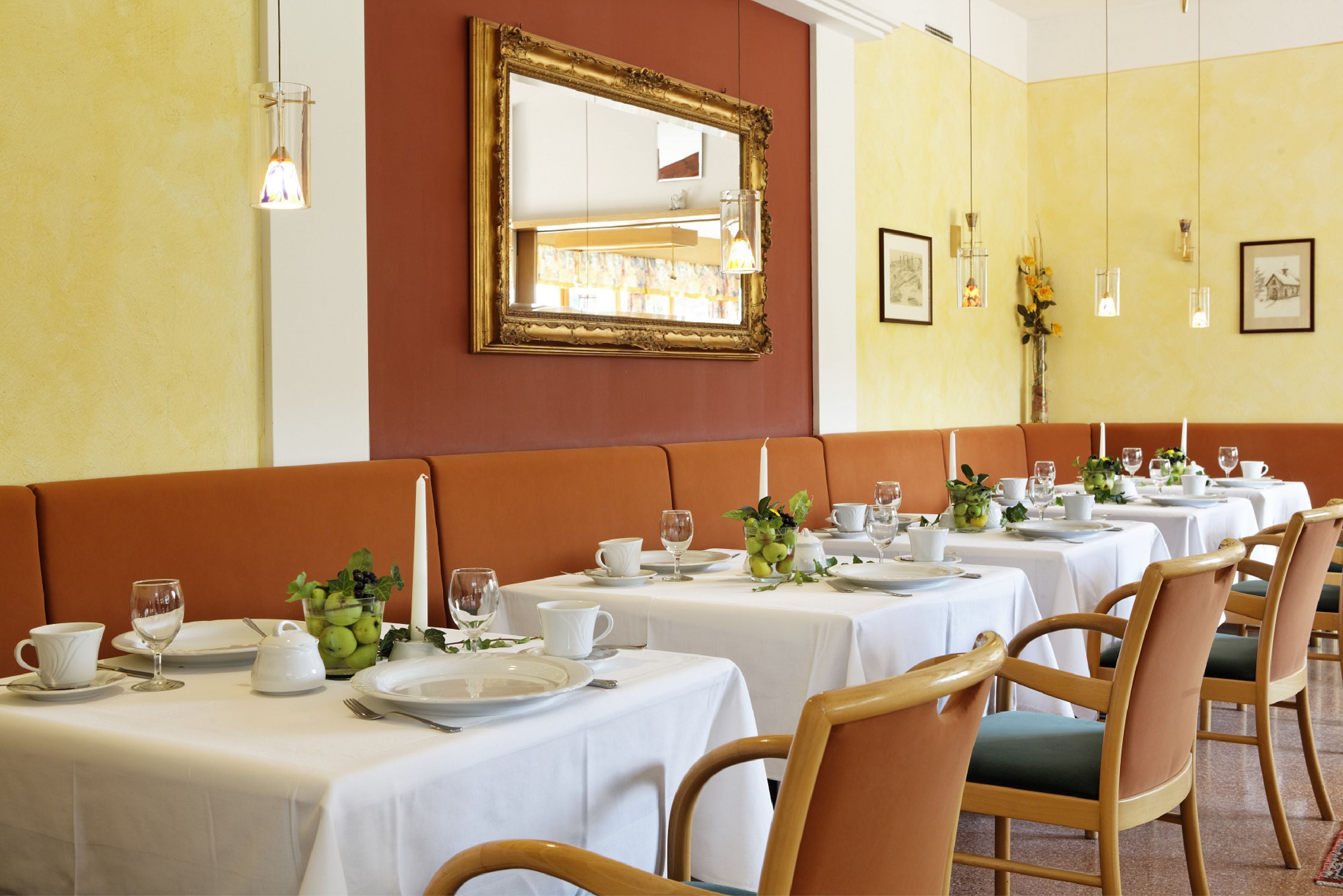 Uma sala de jantar com mesas preparadas para o serviço, com toalhas de mesa brancas, cadeiras de madeira e um espelho grande na parede.