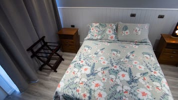Quarto com colcha florida, mesas de cabeceira de madeira, uma cadeira de madeira e uma janela com cortinas.