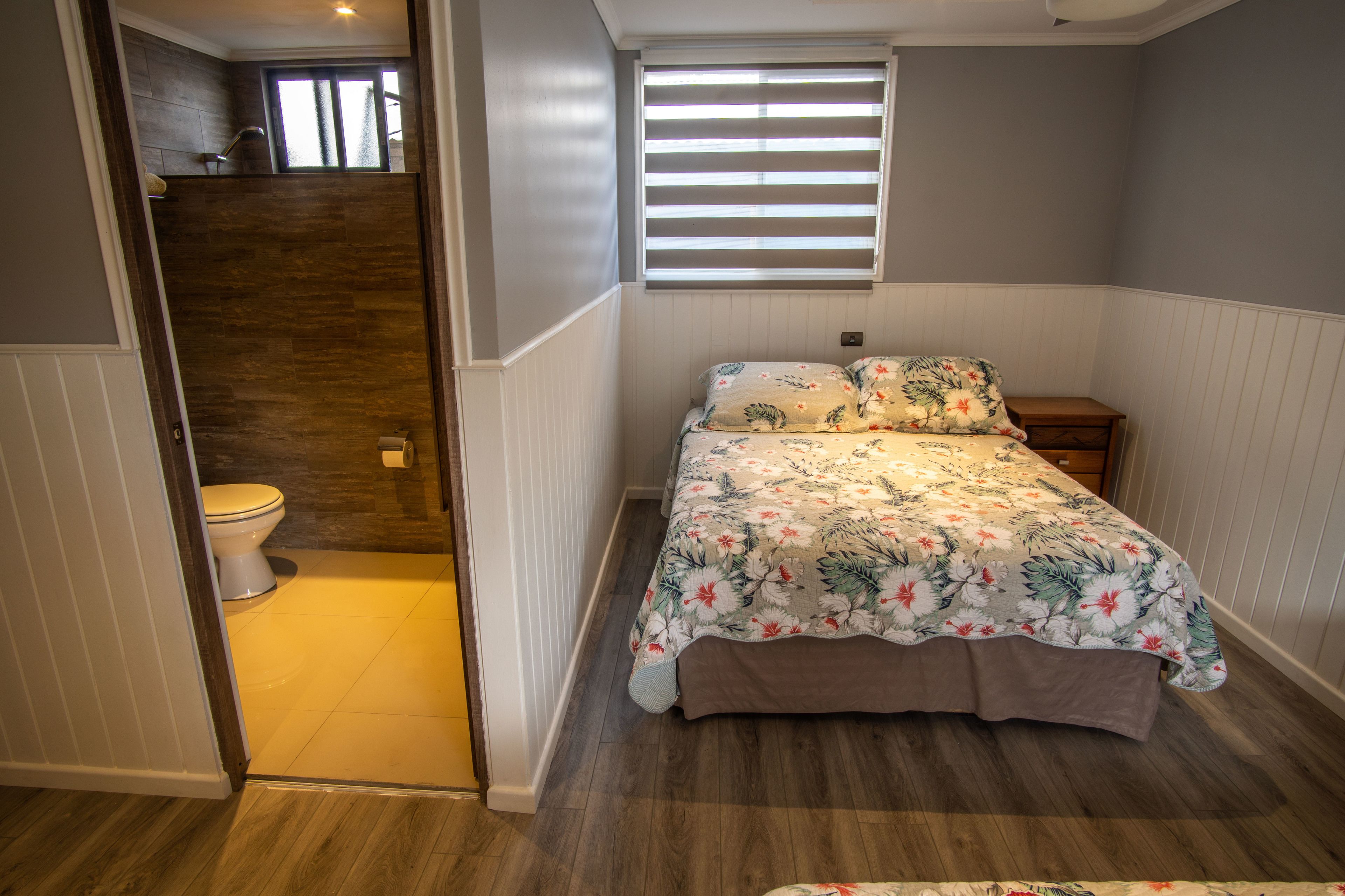 Quarto com cama, janela com persianas, piso de madeira e vaso sanitário.