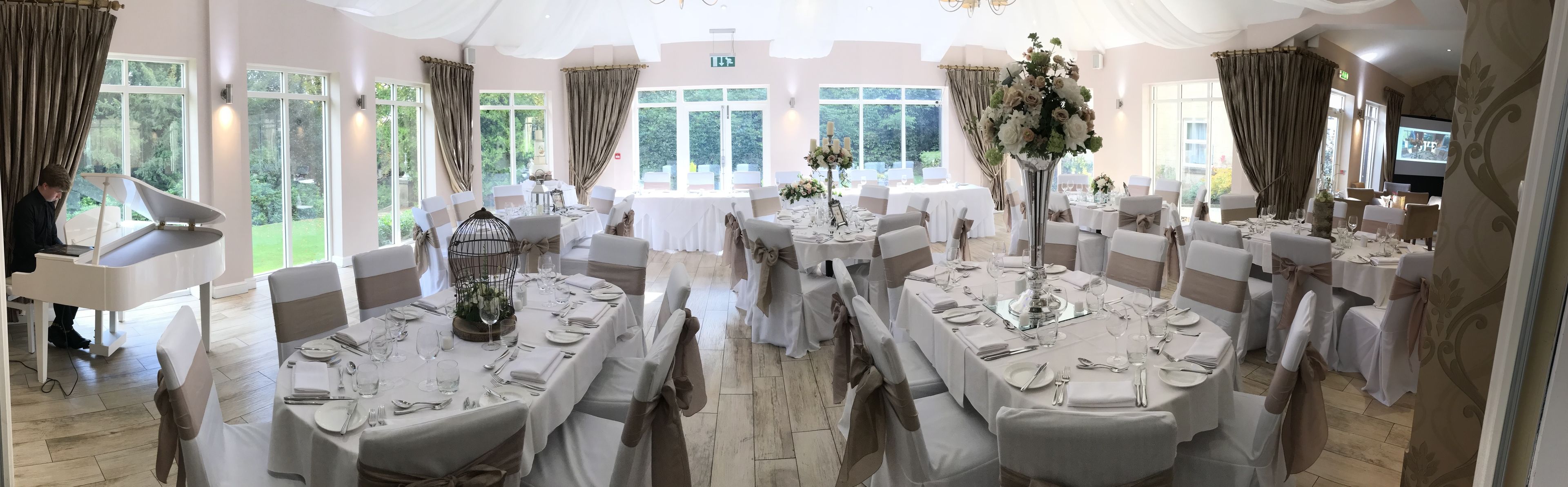 Uma sala de banquetes com várias mesas redondas preparadas para um evento formal, grandes janelas com vista para o exterior e uma tela exibindo um vídeo.