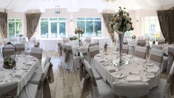 Uma sala de banquetes com várias mesas redondas preparadas para um evento formal, grandes janelas com vista para o exterior e uma tela exibindo um vídeo.