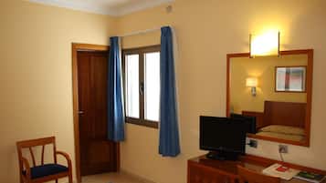 Quarto de hotel com uma escrivaninha de madeira, televisão, cadeira, janela com cortinas e espelho com luminária.