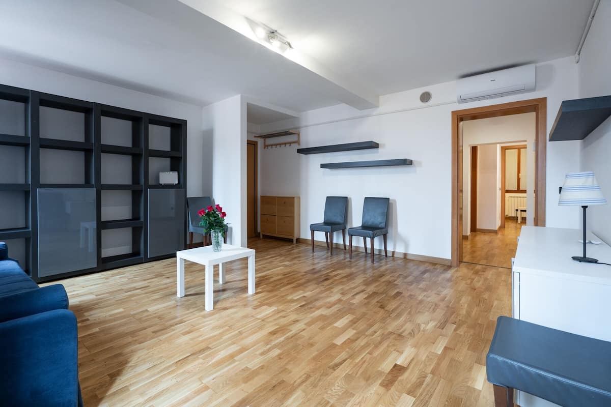 Uma sala de estar espaçosa, com piso de madeira, uma mesa de centro branca, duas cadeiras azuis, uma estante preta e vista para outro cômodo por uma porta aberta.