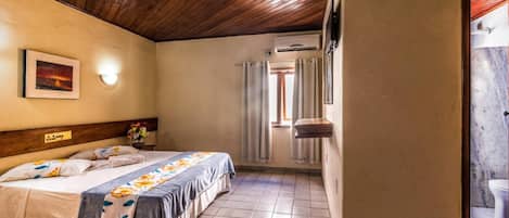 Quarto com teto de madeira, cama com colcha florida, mesa de cabeceira com um vaso, luminária na parede e janela com cortinas.