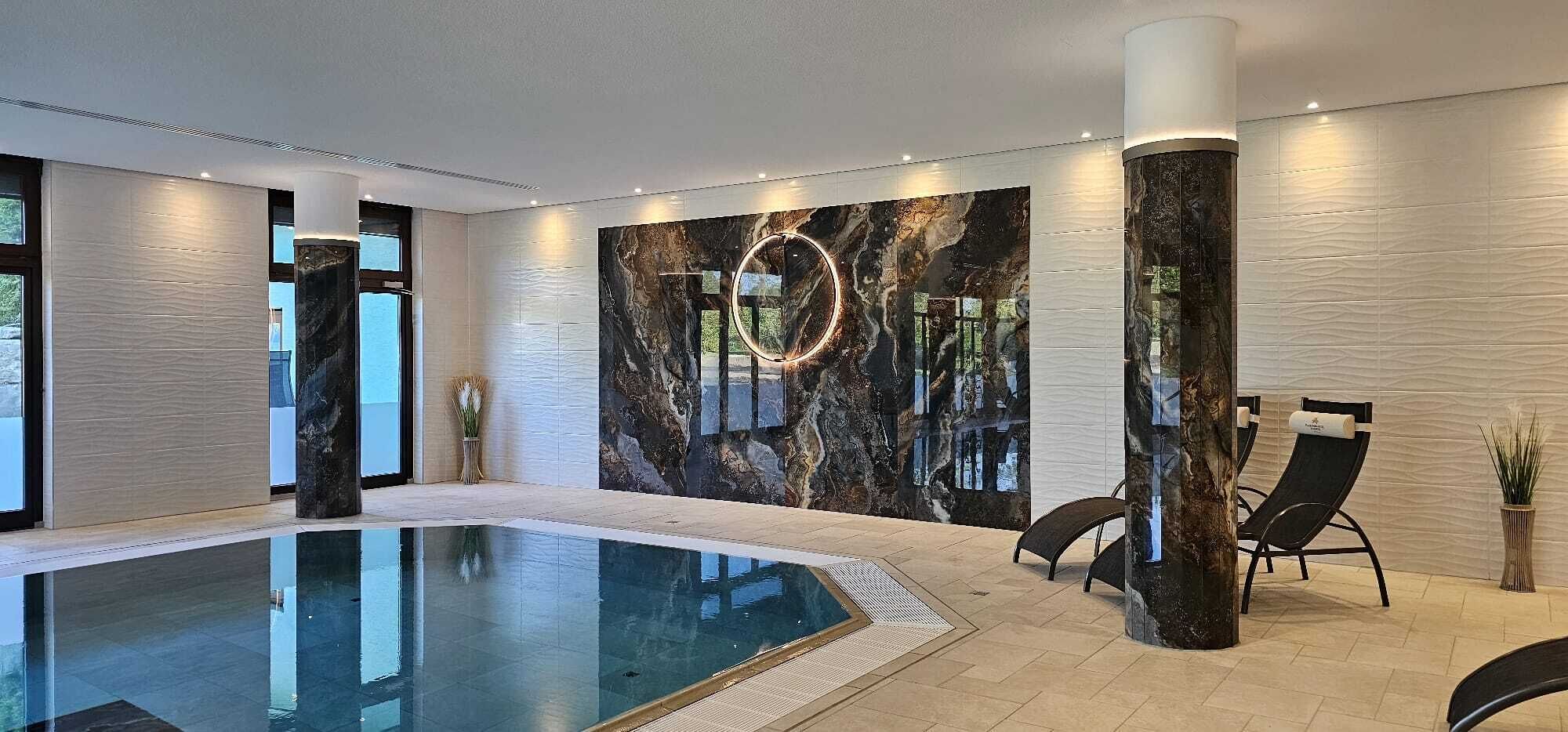 Uma área interna de piscina moderna com uma luminária circular grande na parede, duas espreguiçadeiras e uma piscina transparente.