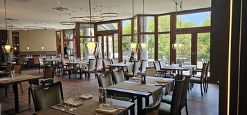 Um restaurante espaçoso com tetos altos, grandes janelas e iluminação pendente moderna. Mesas estão organizadas com cadeiras, e o interior apresenta uma combinação de elementos em madeira e metal.