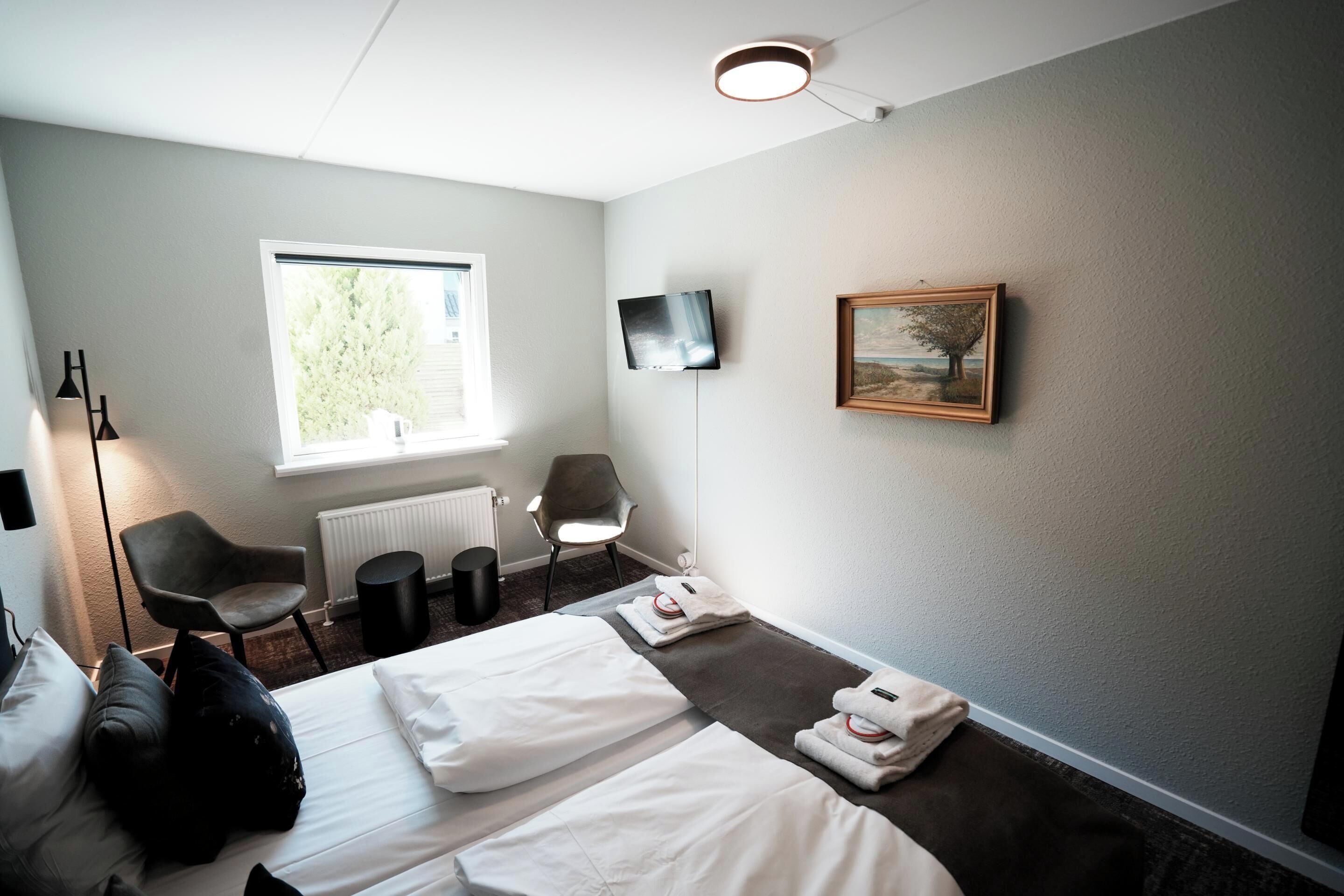 Quarto de hotel com duas camas, uma TV fixada na parede, uma janela com vista para árvores e um quadro na parede.
