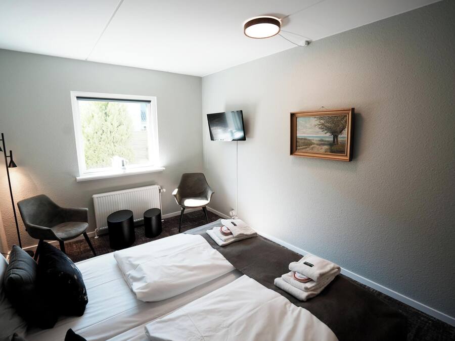 Quarto de hotel com duas camas, uma TV fixada na parede, uma janela com vista para árvores e um quadro na parede.