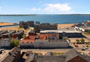 Uma área urbana costeira com mistura de edifícios residenciais e comerciais, uma ponte ao longe e um porto com barcos.