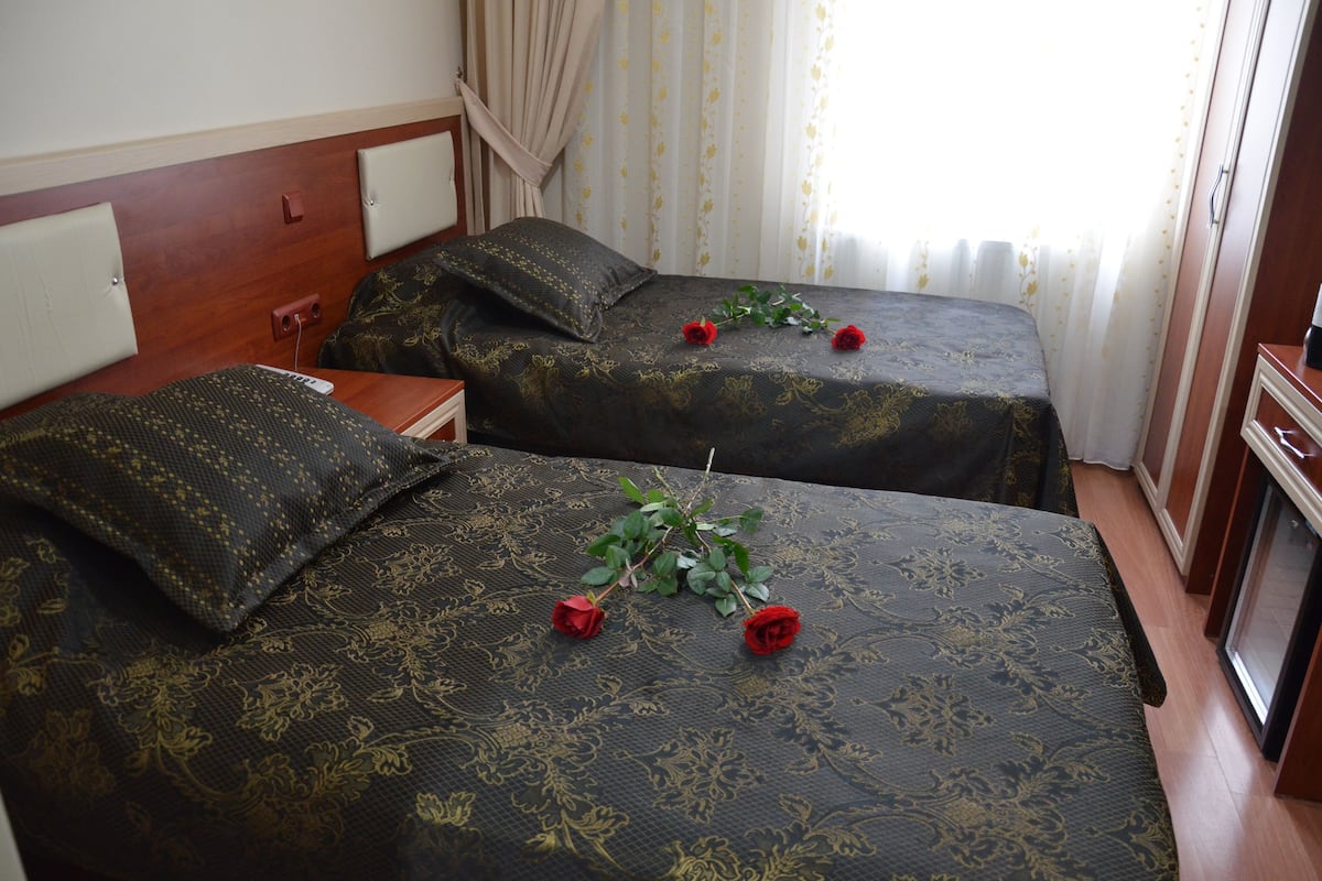 Duas camas com colchas florais e rosas vermelhas, uma cabeceira de madeira e uma janela com cortinas.
