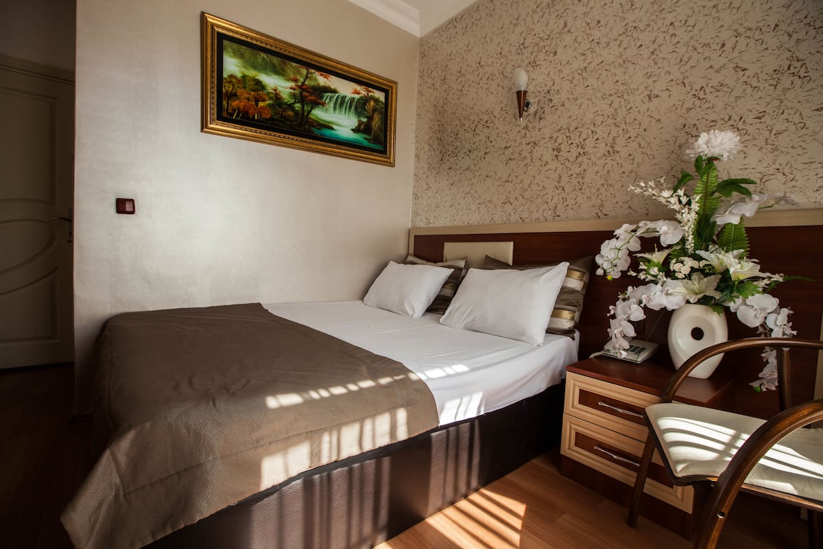 Uma cama bem arrumada com cobertura marrom, uma mesa de cabeceira de madeira com um vaso de flores brancas e um quadro de uma cascata na parede.