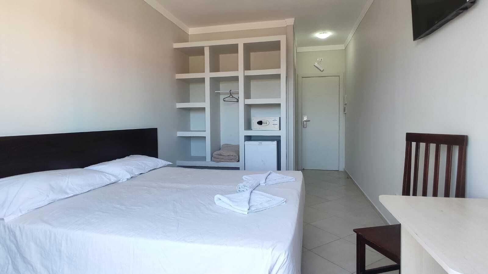 Quarto de hotel com cama, escrivaninha, cadeira, armário e televisão.