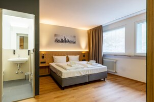 Quarto de hotel moderno com uma cama grande, banheiro com pia e uma pintura de paisagem urbana na parede.