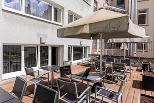Um pátio externo com mesas, cadeiras, um grande guarda-sol e um edifício com janelas e varandas.