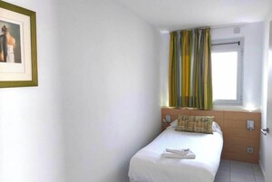 Uma cama de solteiro com roupa de cama branca, um travesseiro e uma toalha dobrada. Há uma janela com cortinas, um interruptor de luz na parede e um quadro emoldurado sobre a cama.