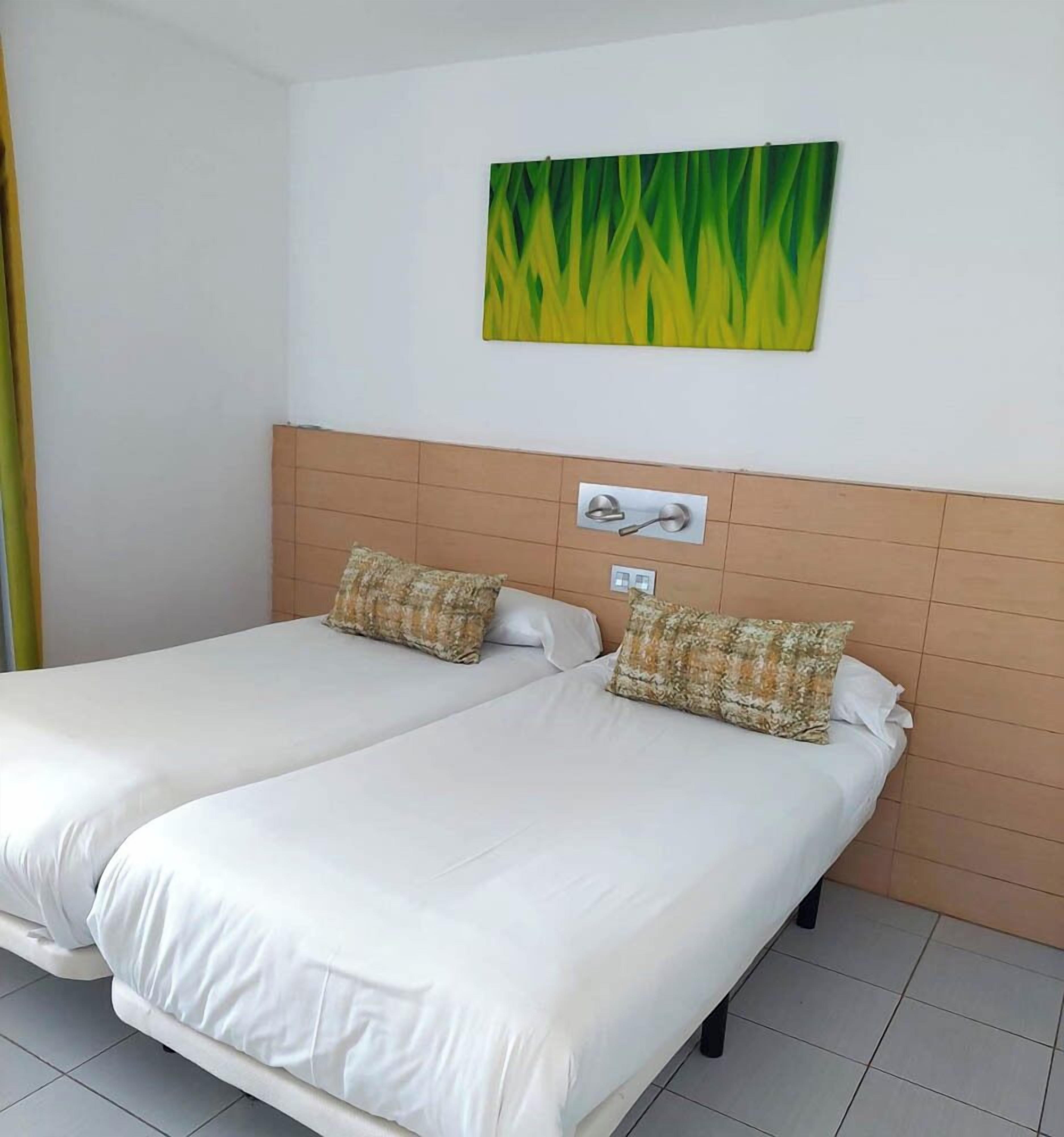 Quarto de hotel com duas camas de solteiro, cabeceira com painel castanho-claro, pintura abstrata verde na parede e janela com cortinas.