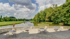 Um pátio com mesas e cadeiras com vista para um lago, rodeado por vegetação exuberante e um céu claro.