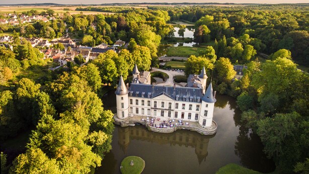 Um edifício grande e histórico com várias torres, rodeado por vegetação exuberante e um corpo de água.
