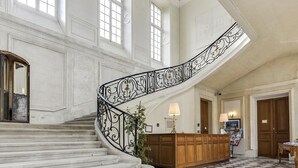 Uma escadaria imponente com grades ornamentadas pretas, um balcão de recepção de madeira e um piso xadrez.