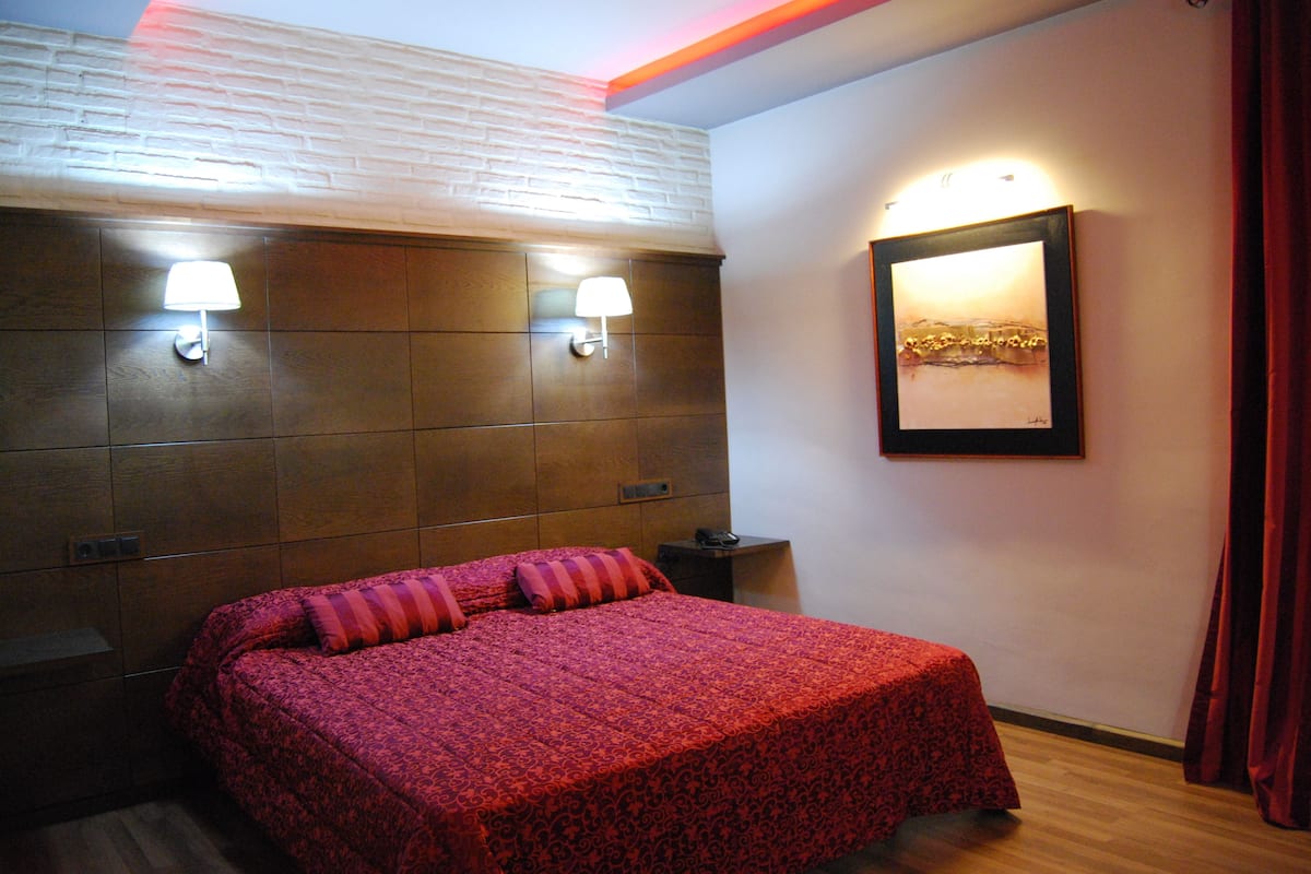 Quarto de hotel com uma cama grande, duas luminárias fixadas na parede, uma obra de arte emoldurada e uma cabeceira de madeira.