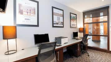 Quarto de hotel com escrivaninha, dois monitores, impressora e pôsteres emoldurados na parede.