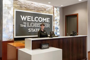 Uma receção com um letreiro de "Welcome to the Lobster State", um letreiro de "Hilton Honors" e um ecrã digital.