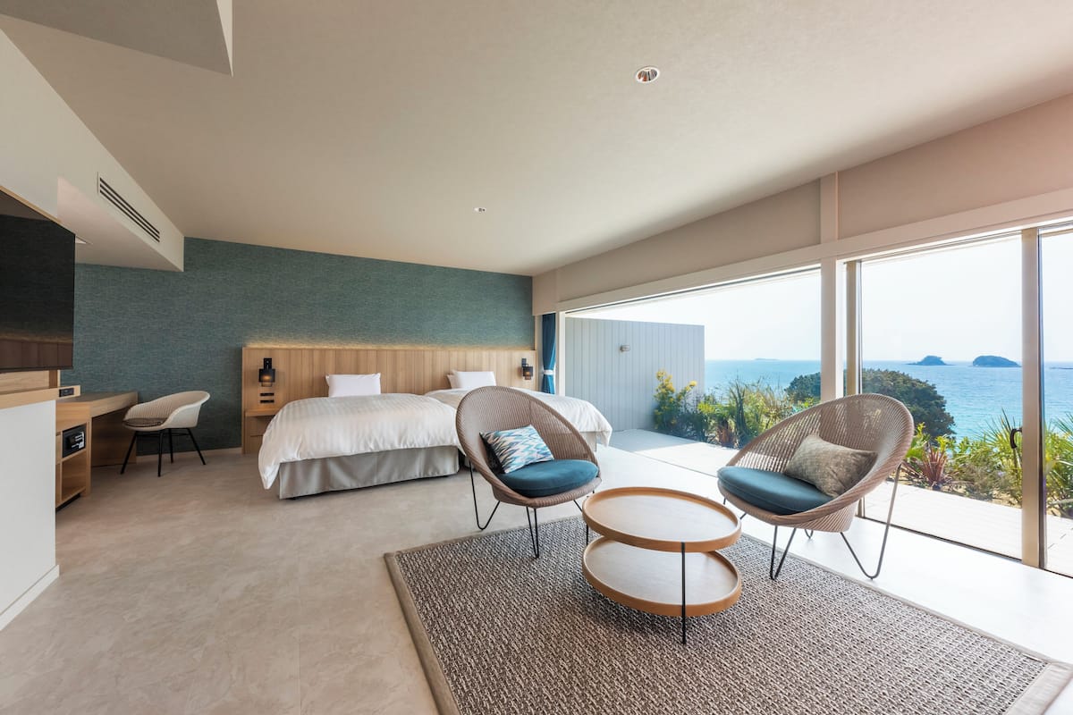 Quarto moderno com cama grande, duas cadeiras, uma escrivaninha e vista para o mar.