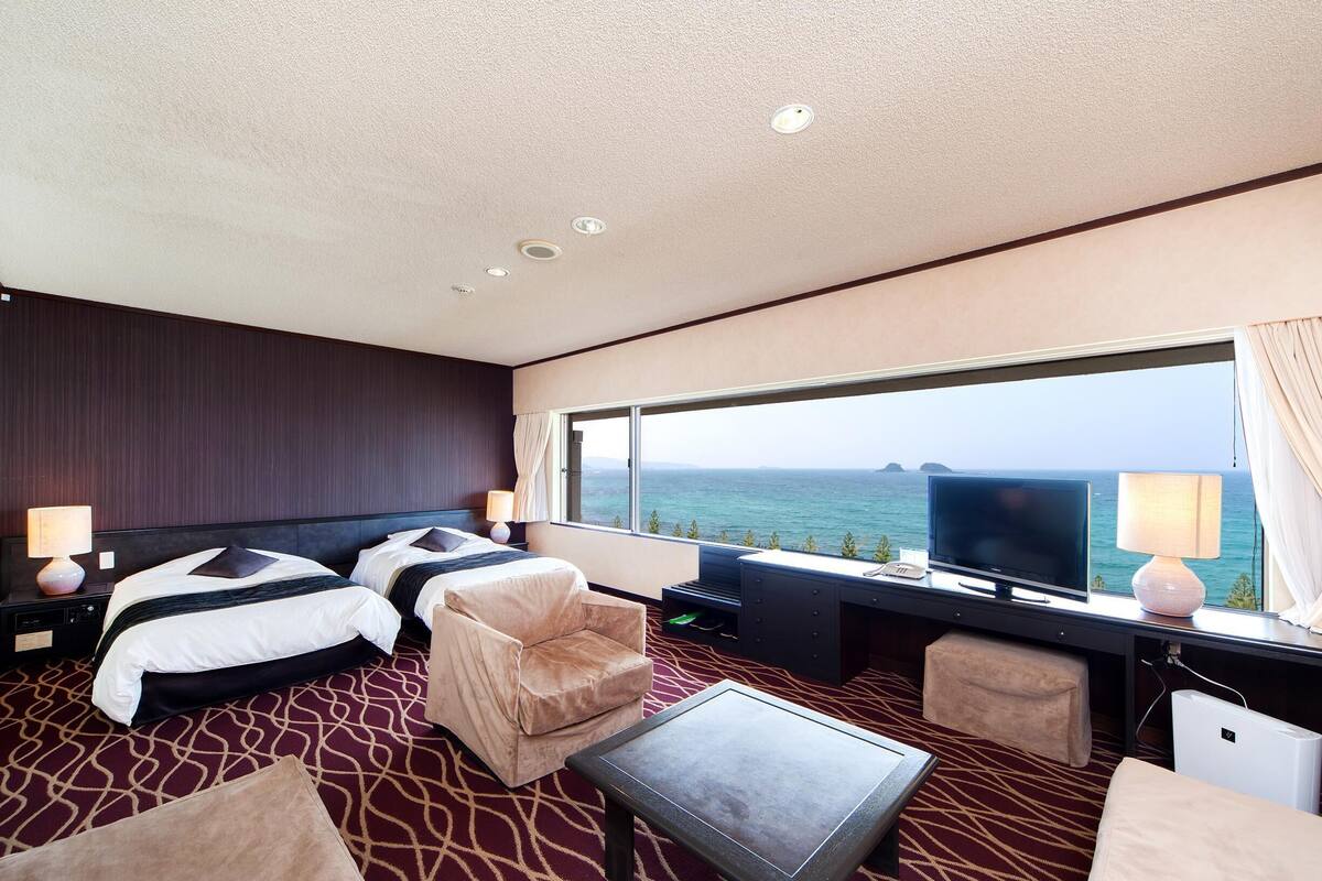 Quarto de hotel com duas camas, uma escrivaninha com televisão, uma mesinha e vista para o mar.