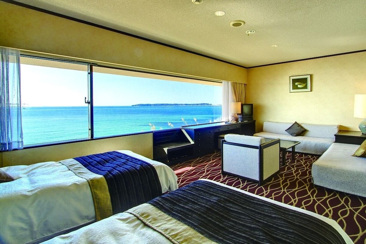 Quarto de hotel com cama, sofá, mesa pequena e televisão. Tem uma janela vista para o oceano.