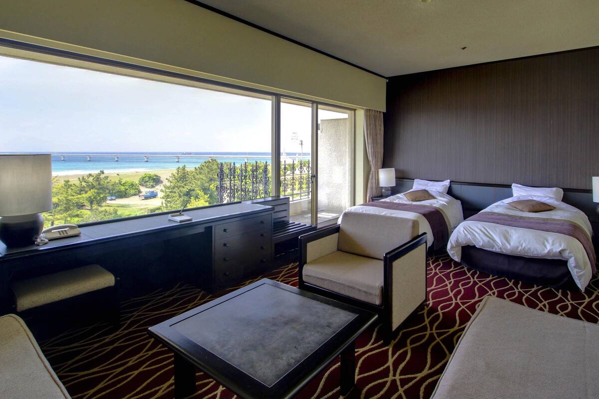 Quarto de hotel com duas camas, uma escrivaninha, uma cadeira e vista para o mar.