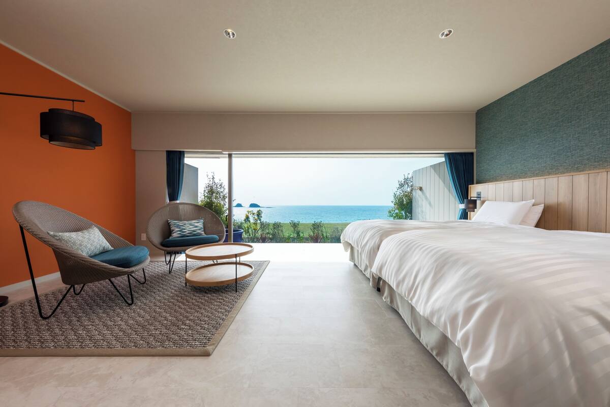 Quarto moderno com cama grande, área de estar com cadeira e mesa, e vista para o mar.