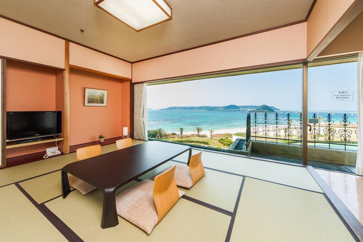 Quarto com uma grande mesa, esteiras de tatame, televisão e vista para o mar.
