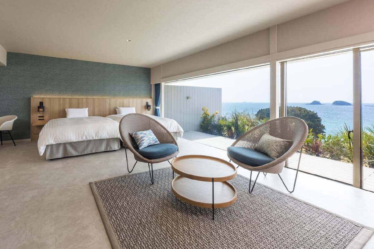 Quarto moderno com uma cama grande, duas cadeiras de vime, uma mesa pequena e vista para o mar.