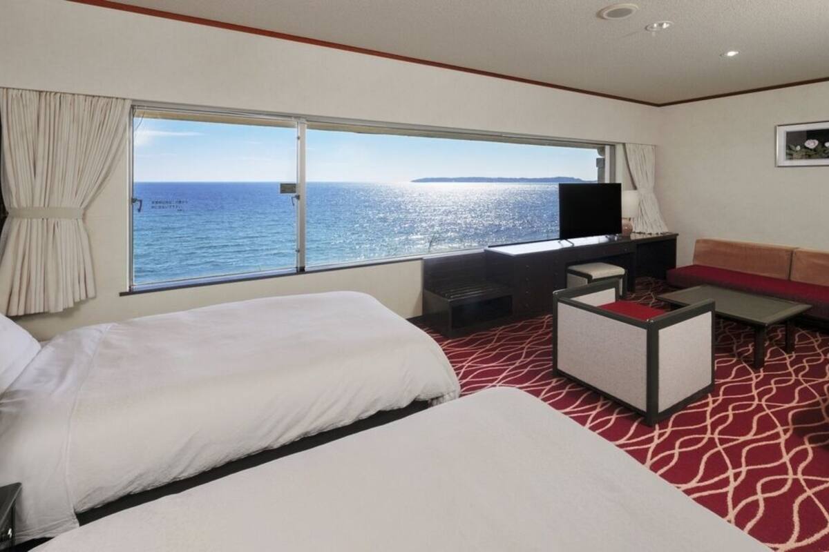 Quarto de hotel com duas camas, uma escrivaninha, televisão, sofá e uma janela grande com vista para o mar.