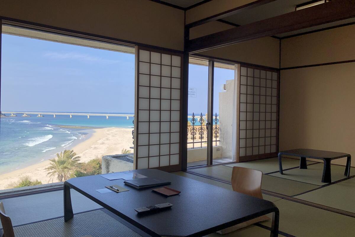 Quarto com piso de tatame, uma mesa baixa com controle remoto e vista para a praia e oceano através de grandes portas deslizantes.