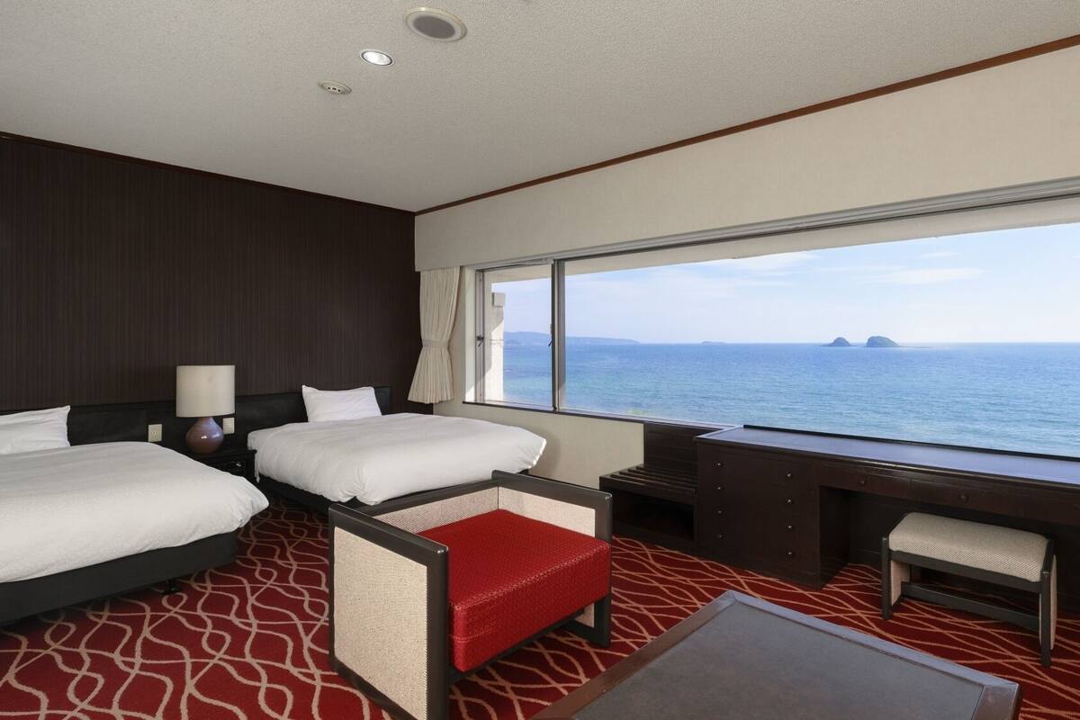 Quarto de hotel com duas camas, uma escrivaninha, uma cadeira e vista para o mar.