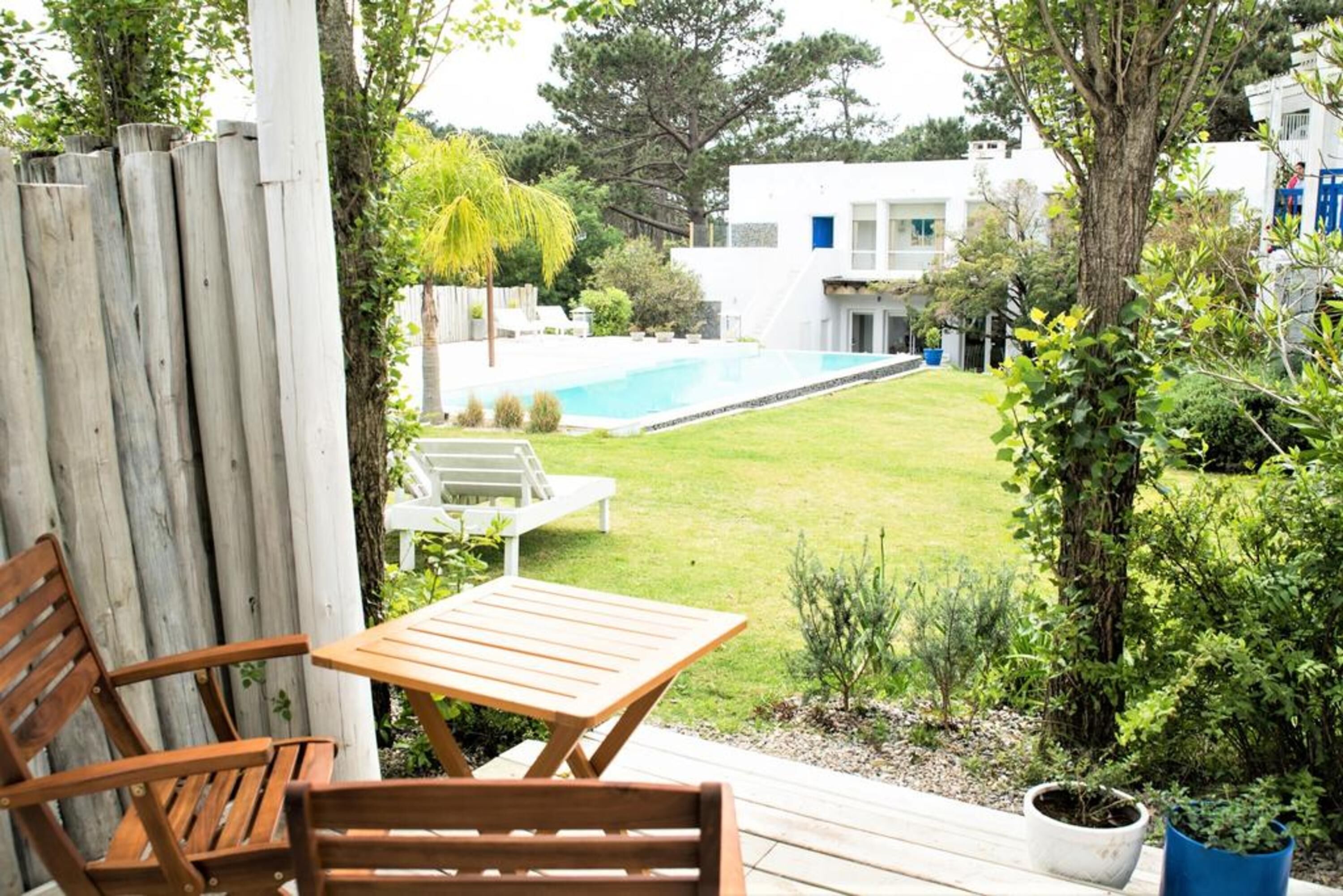 Um deck de madeira com mesa e cadeiras, com vista para uma piscina e um gramado com um edifício branco ao fundo.