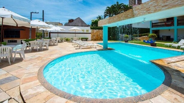 Piscina com espreguiçadeiras e guarda-sóis, cercada por um terraço com piso de pedra.