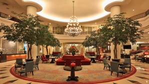 Um hall de entrada com área de estar central, várias mesas e cadeiras, rodeado por árvores grandes e um candelabro.