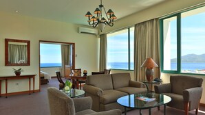 Quarto de hotel com uma grande janela com vista para o mar, um candelabro, um conjunto de sofás e uma mesinha com uma luminária.