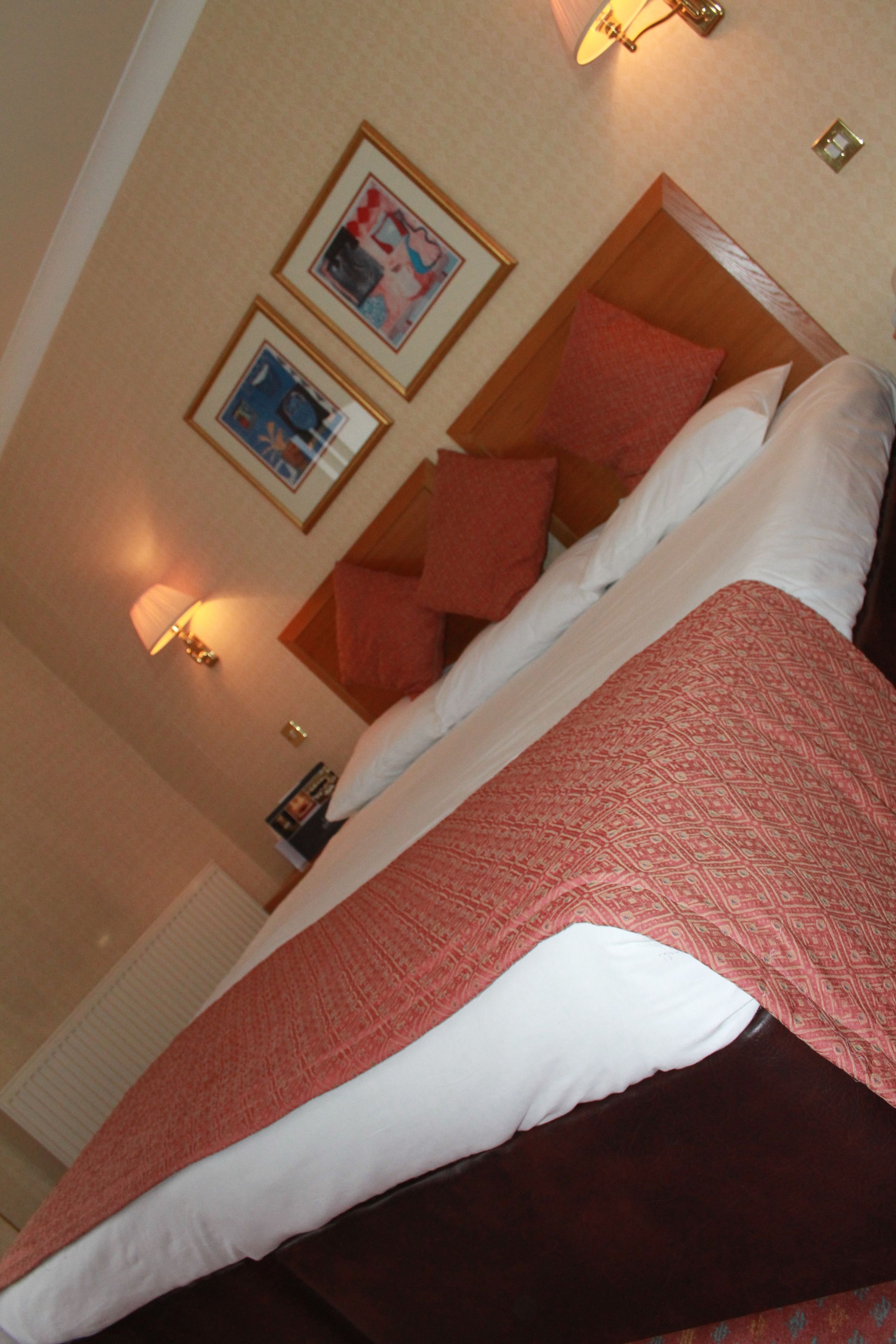 Quarto de hotel com uma cama grande, cabeceira com várias almofadas, luminária na parede e duas quadros emoldurados.