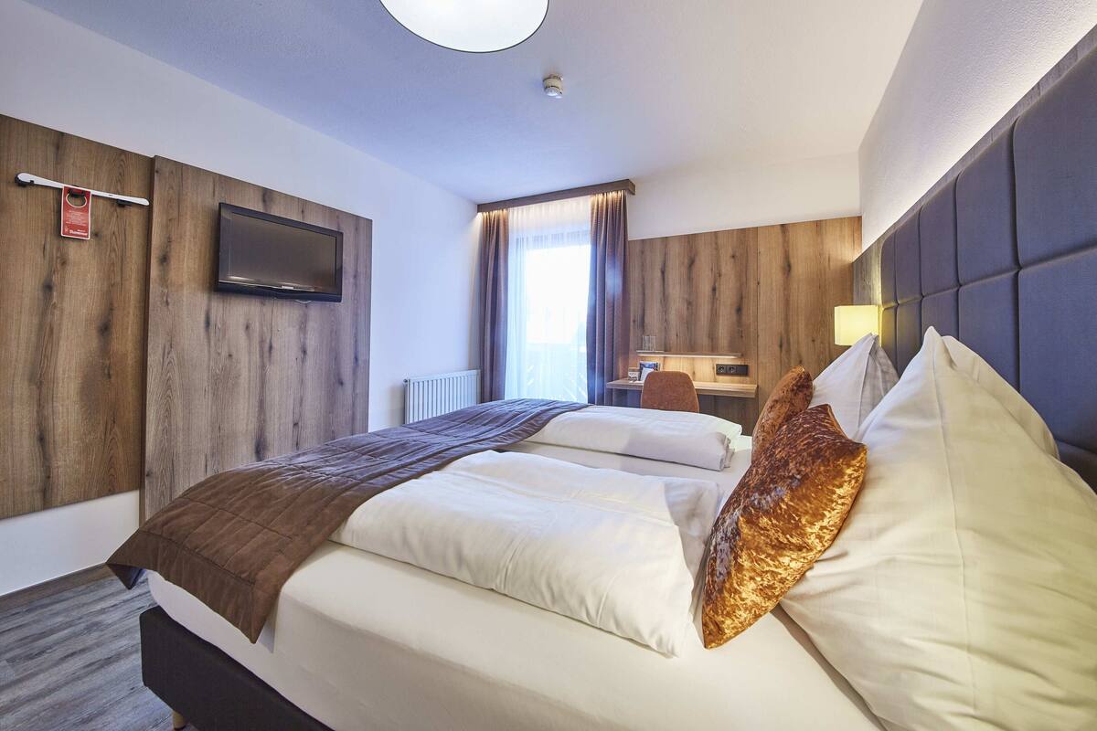 Quarto de hotel com uma cama, uma escrivaninha, uma televisão e uma janela com cortinas.