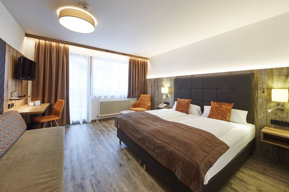Quarto de hotel com uma cama grande, uma escrivaninha com cadeira, televisão na parede e janela com cortinas.