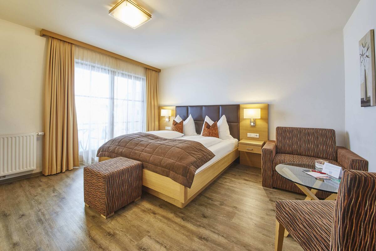 Quarto de hotel com uma cama grande, duas mesas de cabeceira com luminárias, uma pequena mesa redonda com uma cadeira e uma janela com cortinas.
